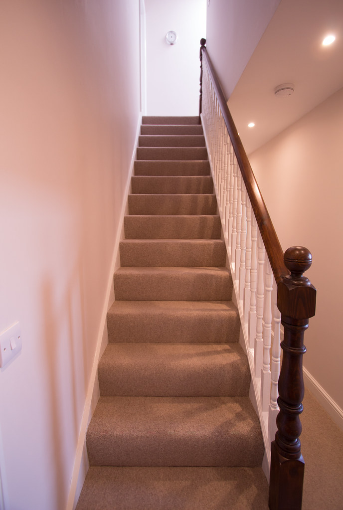 Carpeted interior staircase rising toward a warmly lit landing in a pale-walled corridor, with a white balustrade and dark wood handrail