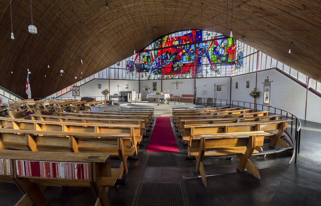St John the Baptist Church interior, Ermine Estate: saddle-shaped timber roof, semicircular pews around the altar, and an abstract stained-glass window