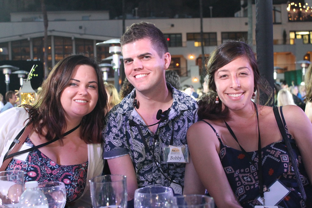 Three attendees share smiles at a casual evening reception, seated around a table with drinks