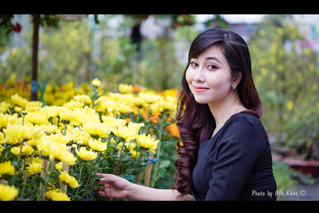 Portrait amid yellow chrysanthemums: a woman in a black dress bathed in soft garden light