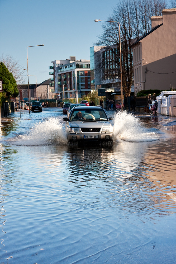 Cork flood scene: silver SUV splashes through a waterlogged city street near the UCC campus