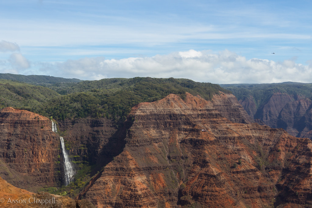 Waimea Canyon, Kauai: Stratified red cliffs, a waterfall, and a lush rim beneath a Hawaiian sky