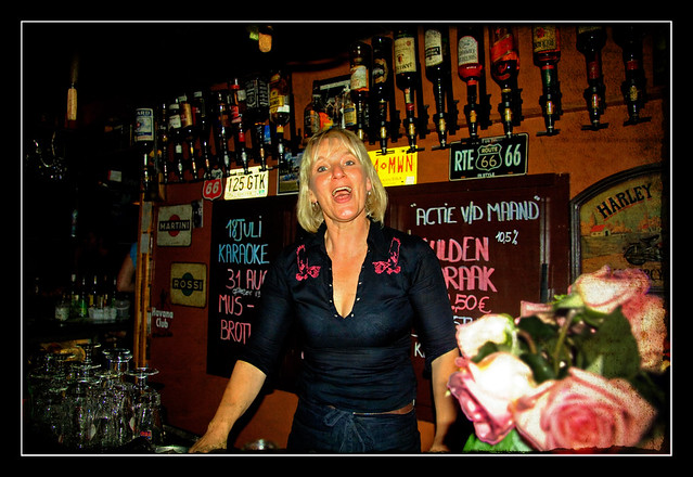 Smiling bartender behind a busy bar during karaoke night, bottles on shelves, chalkboard menus in the background, and a bouquet visible in the foreground.