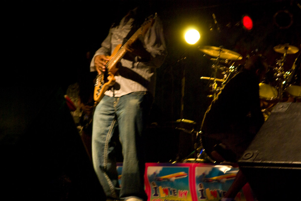 Live reggae-style performance: guitarist in the foreground with drummer visible on stage