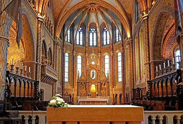 Interior of Matthias Church in Budapest, view down the nave toward the ornate main altar with tall stained-glass windows, ribbed vaults, and dark wooden choir stalls.