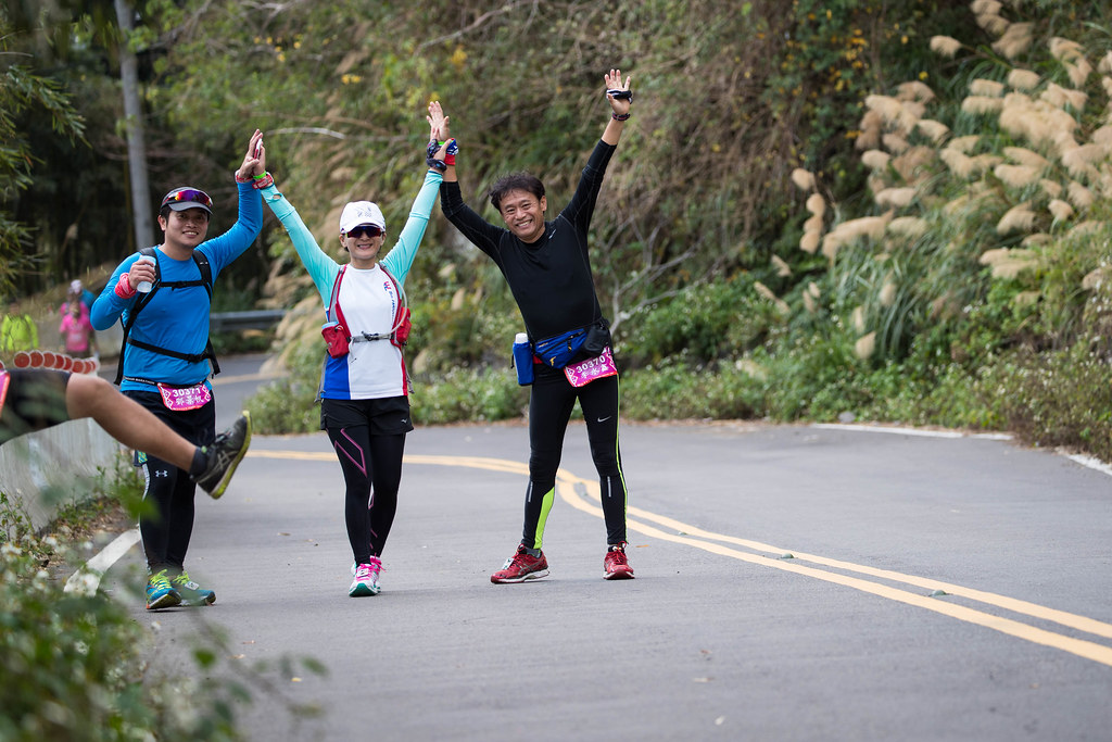 Three runners celebrate with raised arms on a tree-lined road during a daytime race