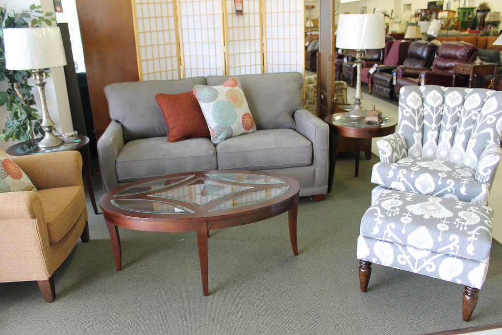Showroom scene featuring a gray sofa with orange and patterned pillows, flanked by beige and damask-patterned chairs around a glass-top coffee table