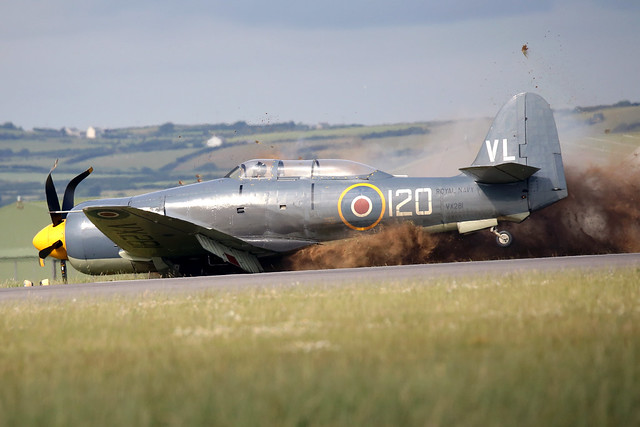 Sea Fury on take-off roll with wheels up; silver fuselage bears Royal Navy roundel and 120, VL tail code; dust plume rises from the runway as the propeller blades blur.