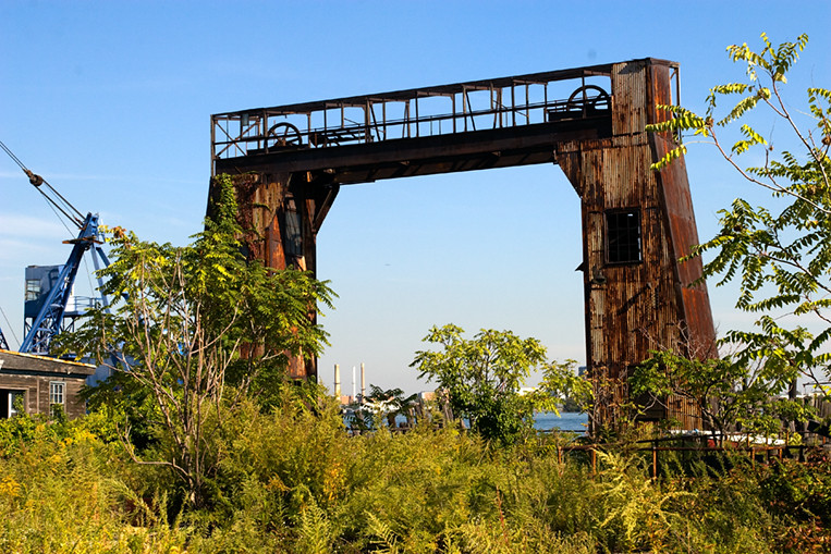 Rusting ferry gantry on North Brother Island shoreline, framed by overgrown vegetation and distant industrial skyline