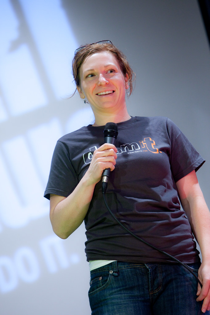Youth Tech Event: Female presenter in a gray tee speaks into a handheld microphone on stage.