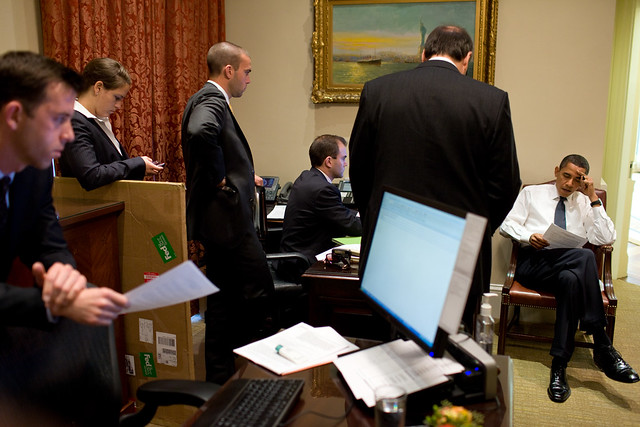 Aides in dark suits gather around a central desk with a computer monitor in a White House office; a man in a white shirt and tie sits nearby, reading a document.