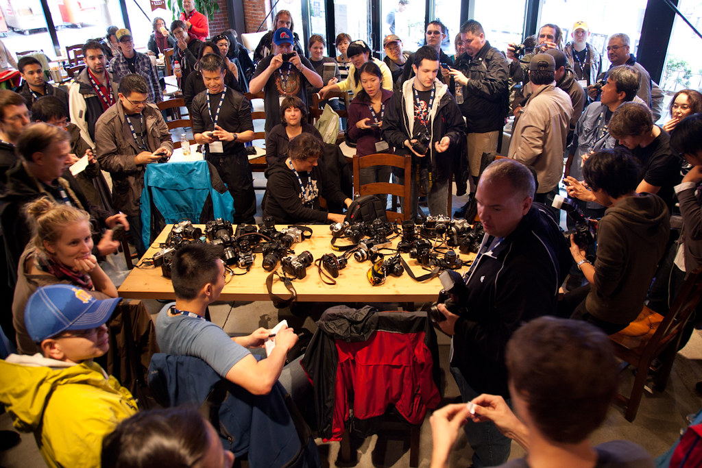 Indoor camera-gear meetup: photographers circle a long table of vintage and modern cameras, lenses, and accessories
