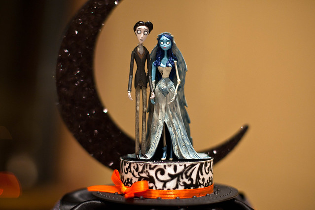 Corpse Bride and Groom figurines on a patterned cake stand with an orange ribbon, set against a crescent-moon backdrop and softly blurred warm background.