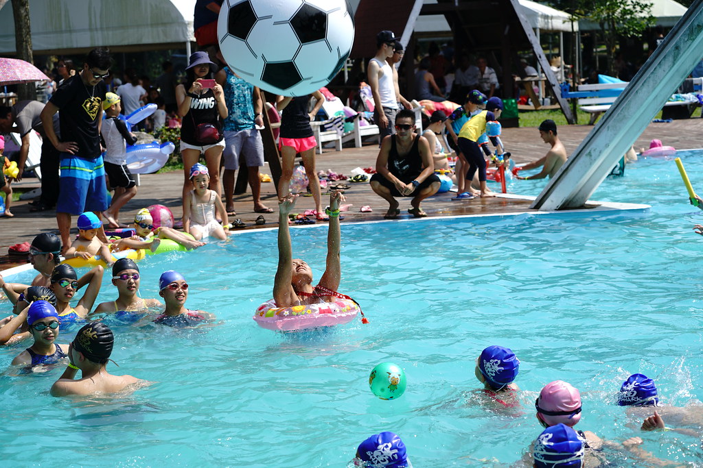 Child on inflatable ring reaches for a large floating soccer ball at a crowded, sunlit public pool.