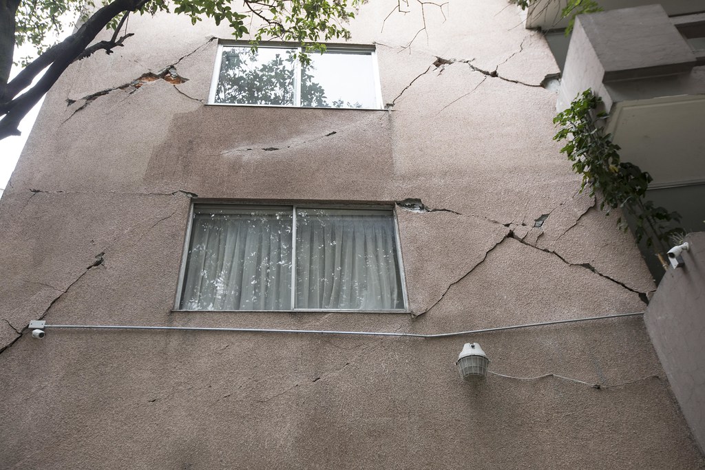 Cracked pink stucco facade of a multi-story building with two windows, sheer curtains, and exposed electrical conduits