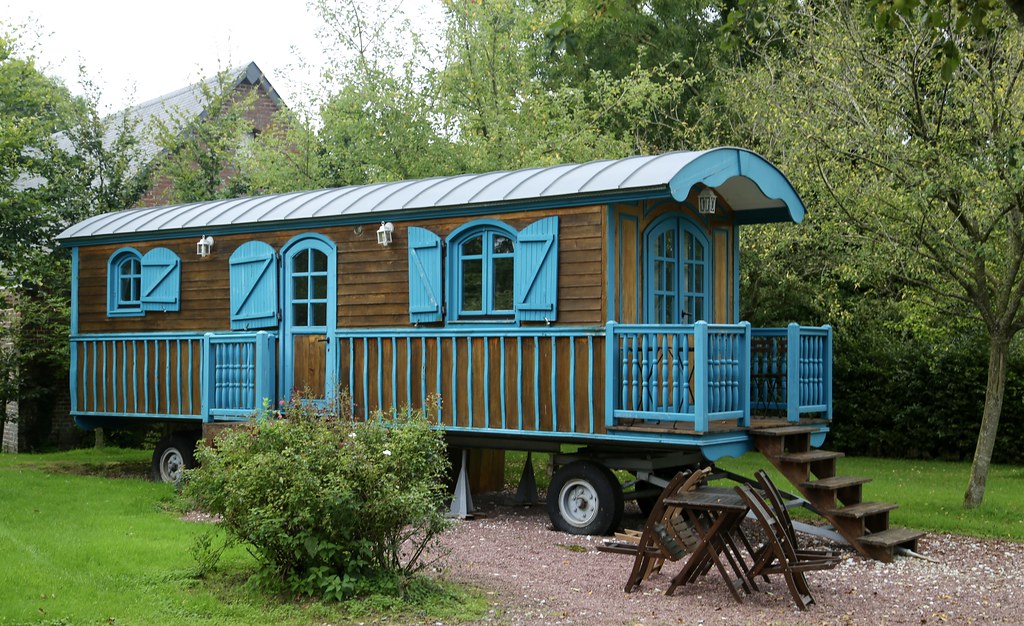 Blue wooden caravan on a wheeled trailer with turquoise trim and wraparound deck in a lush garden