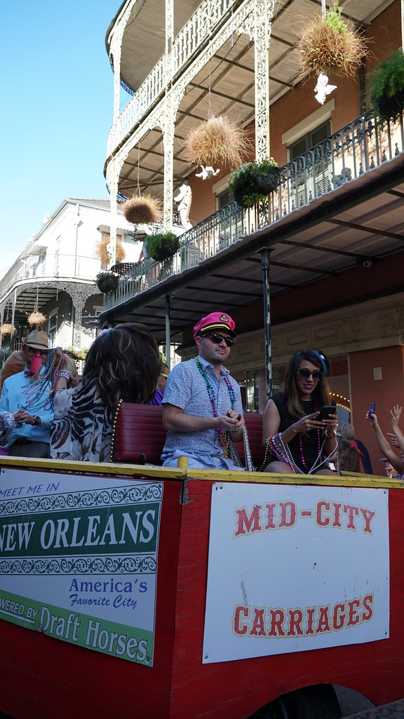 New Orleans Parade Float with Beaded Revelers Under Historic Iron Balconies (Mid-City Carriages Signage)