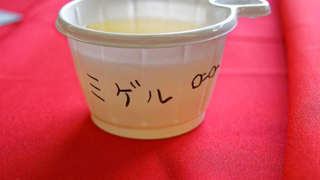 Miguel label (ミゲル) on a white plastic cup with pale yellow liquid against a red fabric backdrop
