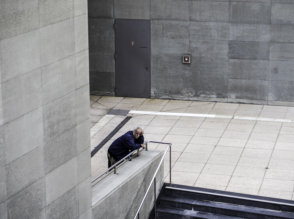Adult man leaning on a metal railing in a minimalist concrete stairwell framed by stark geometric architecture