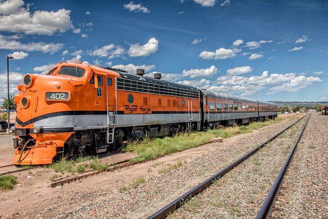 Orange Royal Gorge locomotive 402 with vintage passenger cars on a gravel rail yard, rails receding toward the horizon under a bright blue sky.