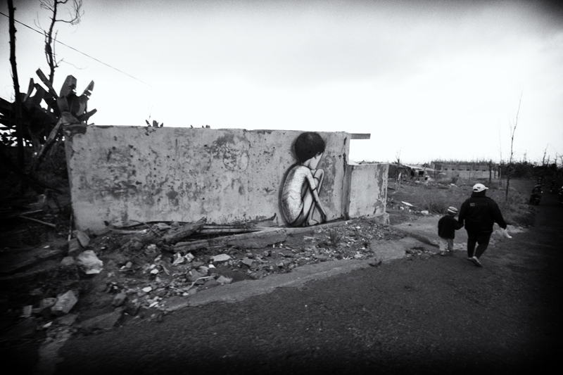 Urban Ruin: Graffiti of a Crouching Figure on a Weathered Wall as an Adult and Child Walk Down a Rubble-Strewn Street