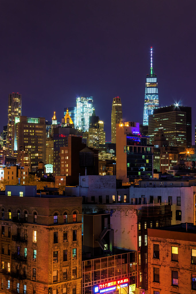 Nighttime panorama of Lower Manhattan: One World Trade Center rises above a mosaic of lit brick buildings
