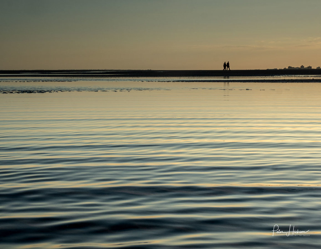 Two silhouettes on Hayling Island's low-tide shoreline as rippled water glows at dusk