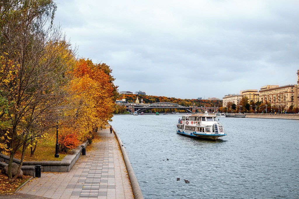 Autumn riverside promenade in Moscow: a passenger boat gliding along the Moskva River past a tree-lined embankment and distant bridge