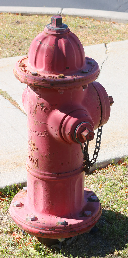 Close-up of a weathered red fire hydrant on a sunlit sidewalk, showing rust, chipped paint, and a diagonally draped outlet chain