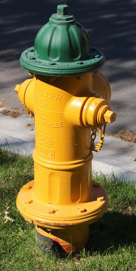 Yellow cast-iron fire hydrant with a green domed cap on a sunlit grassy verge beside the sidewalk