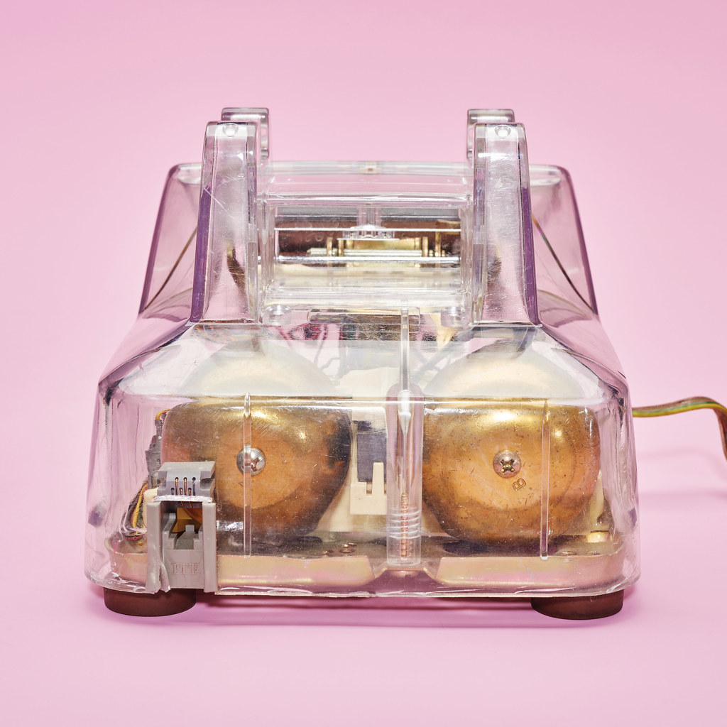 Transparent vintage desk telephone with exposed brass bells against a pink studio backdrop