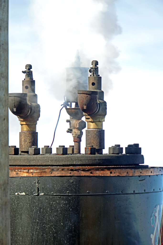 Close-up of weathered boiler valves with rising steam above a rusted riveted tank