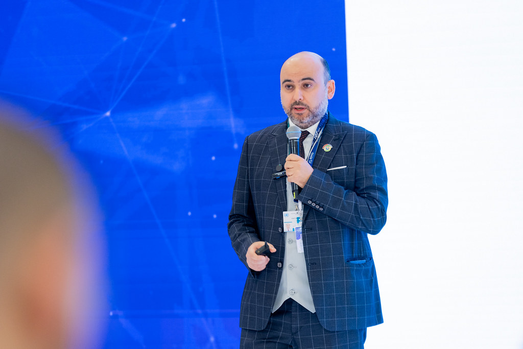 IGF 2024 Day 4, Riyadh: Policy speaker presents on digital governance on a blue, network-inspired stage.
