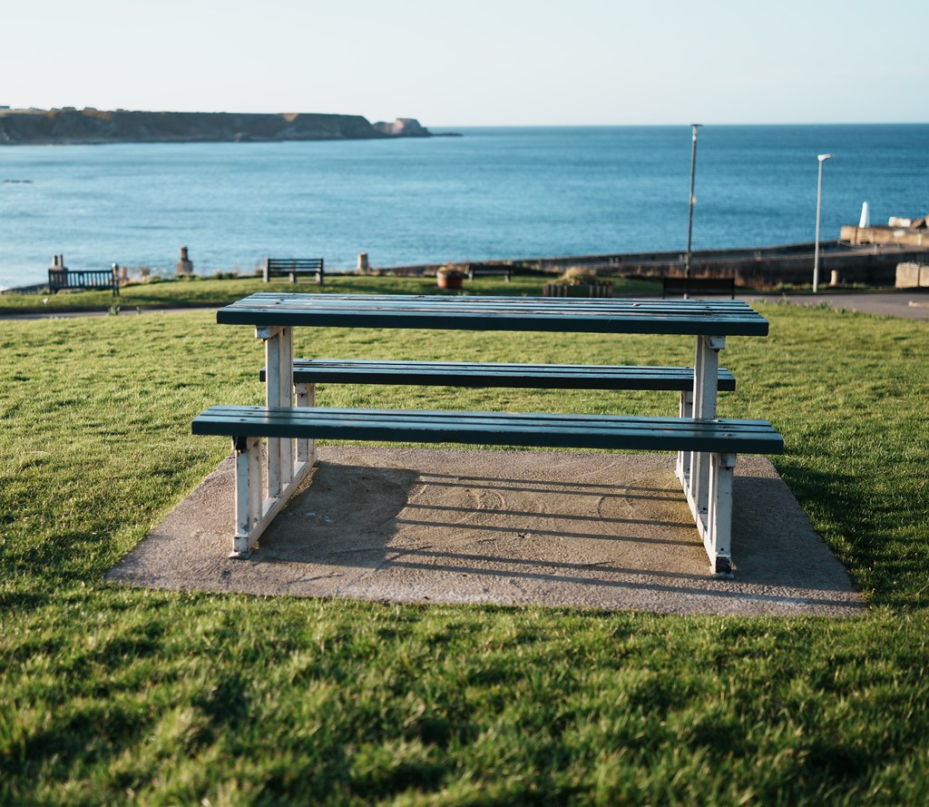 Sunlit coastal park bench by the sea: weathered picnic table overlooking calm blue waters