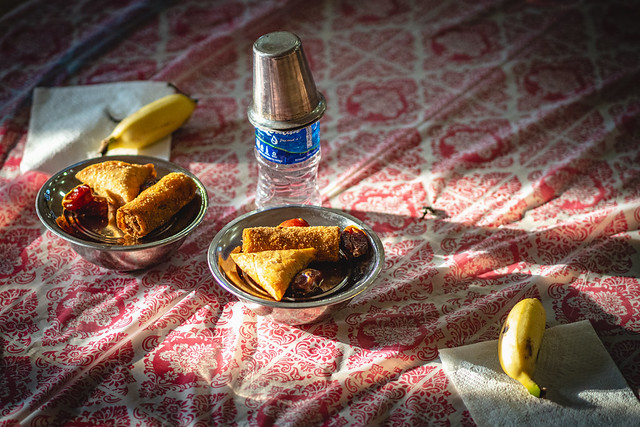 Two metal bowls with pastries and dates on a red patterned cloth, alongside a water bottle, a metal cup, and a banana on a napkin, lit by soft side light.