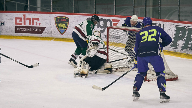 Indoor ice hockey action near the net: goalie in white pads defends while a purple jersey player #32 approaches and a green-and-white skater pressures the crease; sponsor boards and glass visible in bright arena lighting.
