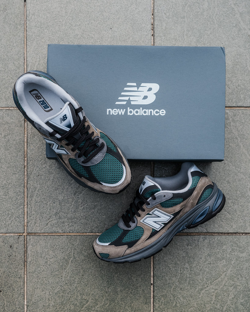 New Balance sneakers with teal mesh and gray suede beside a blue NB box on gray tiled pavement