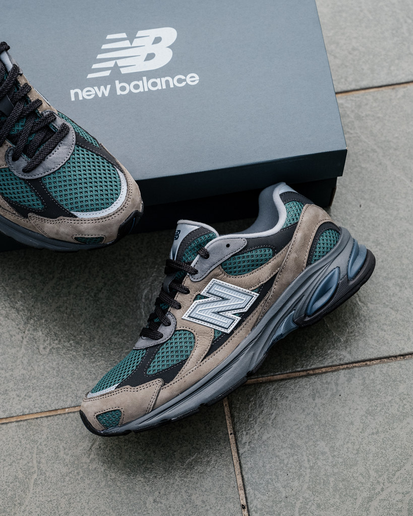New Balance sneakers in teal mesh and gray suede beside an NB shoebox on textured concrete tiles