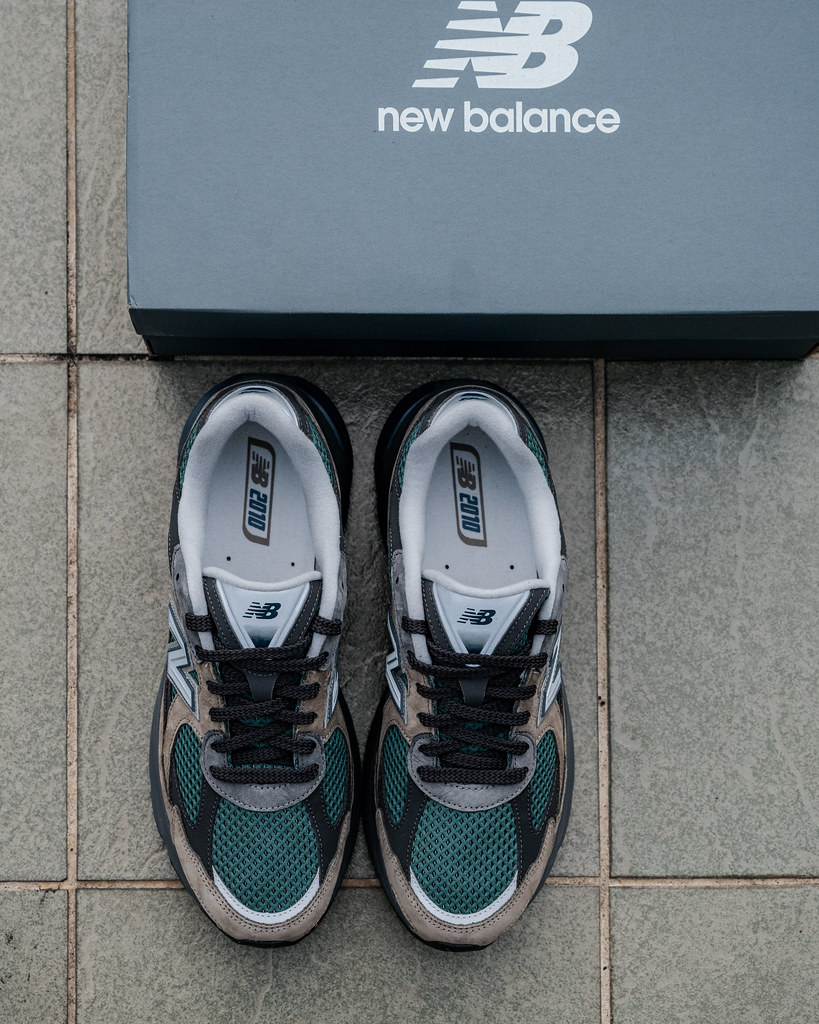 New Balance 2010 sneakers in teal mesh and gray overlays, paired beside a gray NB box on tiled pavement.