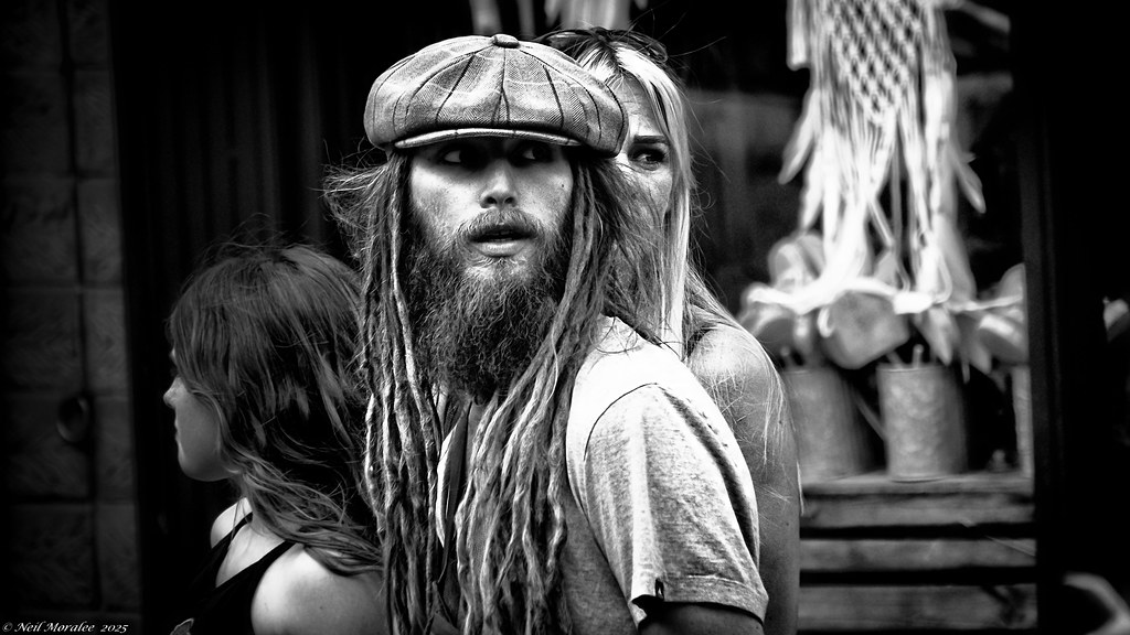 Monochrome street portrait of a bearded man with dreadlocks and a flat cap, surrounded by passersby