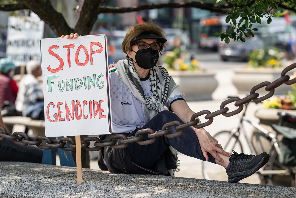 Seated protester with STOP FUNDING GENOCIDE sign and diagonal chain barrier on Chicago's Michigan Avenue Gaza solidarity demonstration