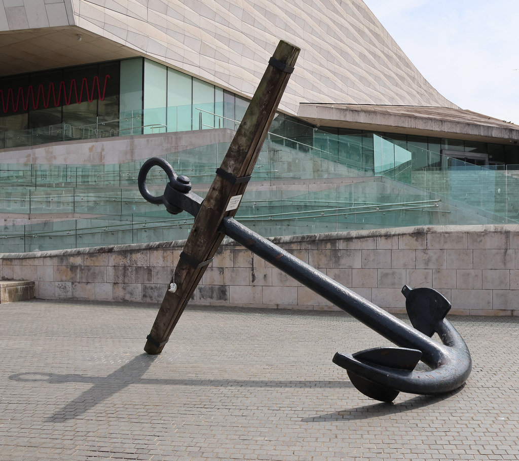 Massive maritime anchor sculpture in a plaza with modern glass architecture backdrop