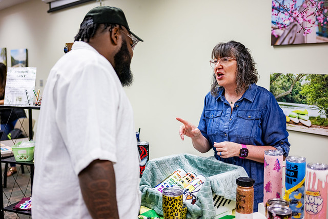 Bearded man in a white shirt talks with a woman in a denim shirt at a Match & Market Carnival booth; table displays colorful tumblers and a fabric bin.
