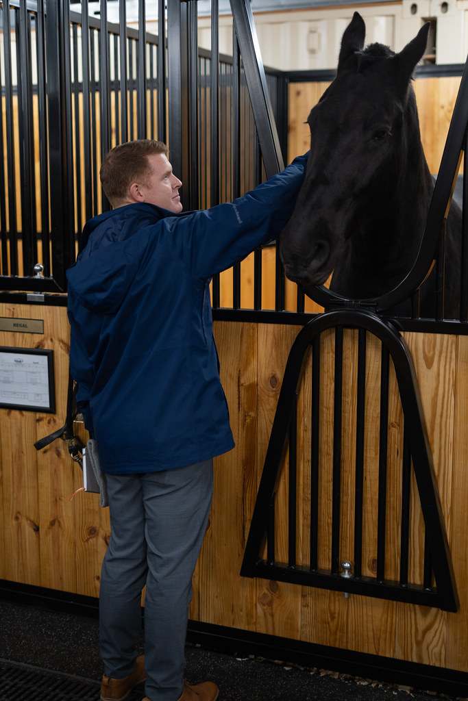 Man in blue jacket strokes a black horse in a wooden stable stall
