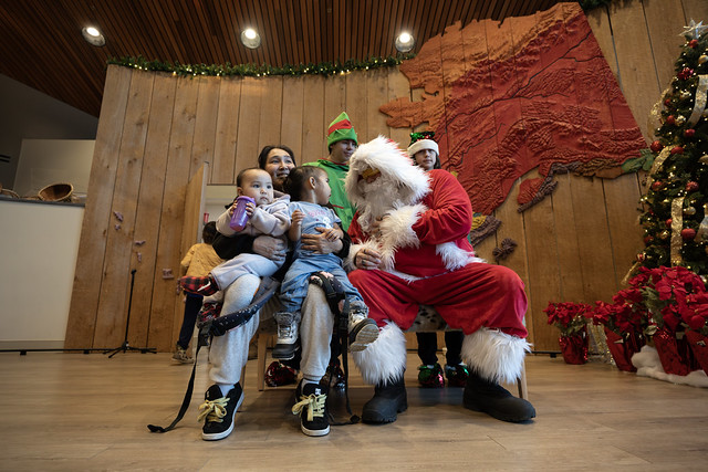 Santa in a red suit with white fur trim sits with children on his lap as volunteers look on, in front of a carved Alaska map wall and a decorated Christmas tree.