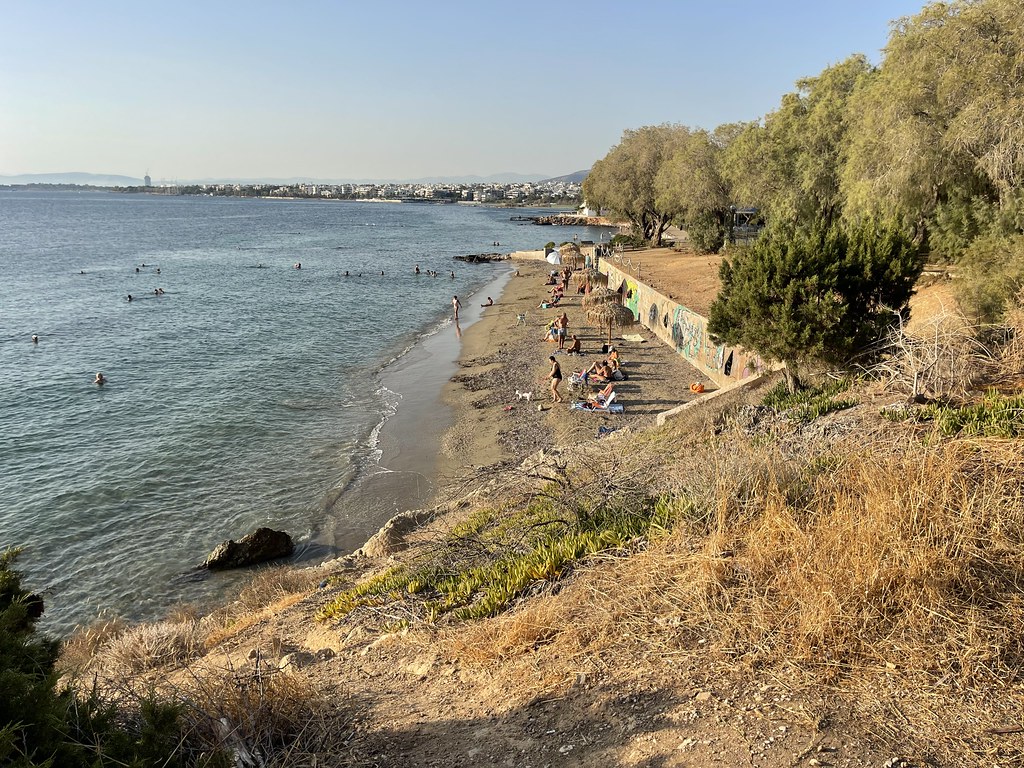 Kouvari Beach, Vouliagmeni, Athens Riviera – Sunlit shoreline with swimmers and a graffiti-lined seawall