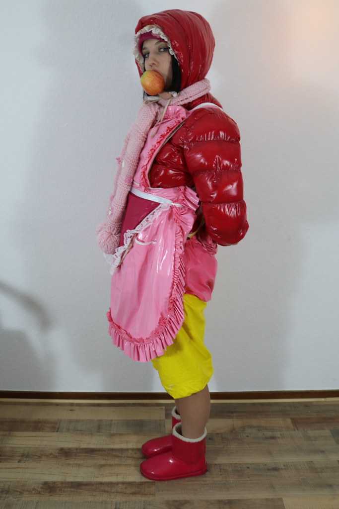 Playful full-body portrait: pink glossy apron over a red puffer jacket, with yellow pants and red boots, holding an apple in the mouth