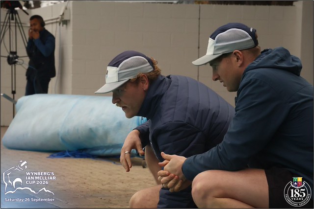 Two players in navy jackets kneel on a pool deck during IMCUP26 warm-up, with a rolled blue mat and a camera operator in the background; event logos appear in the corners.