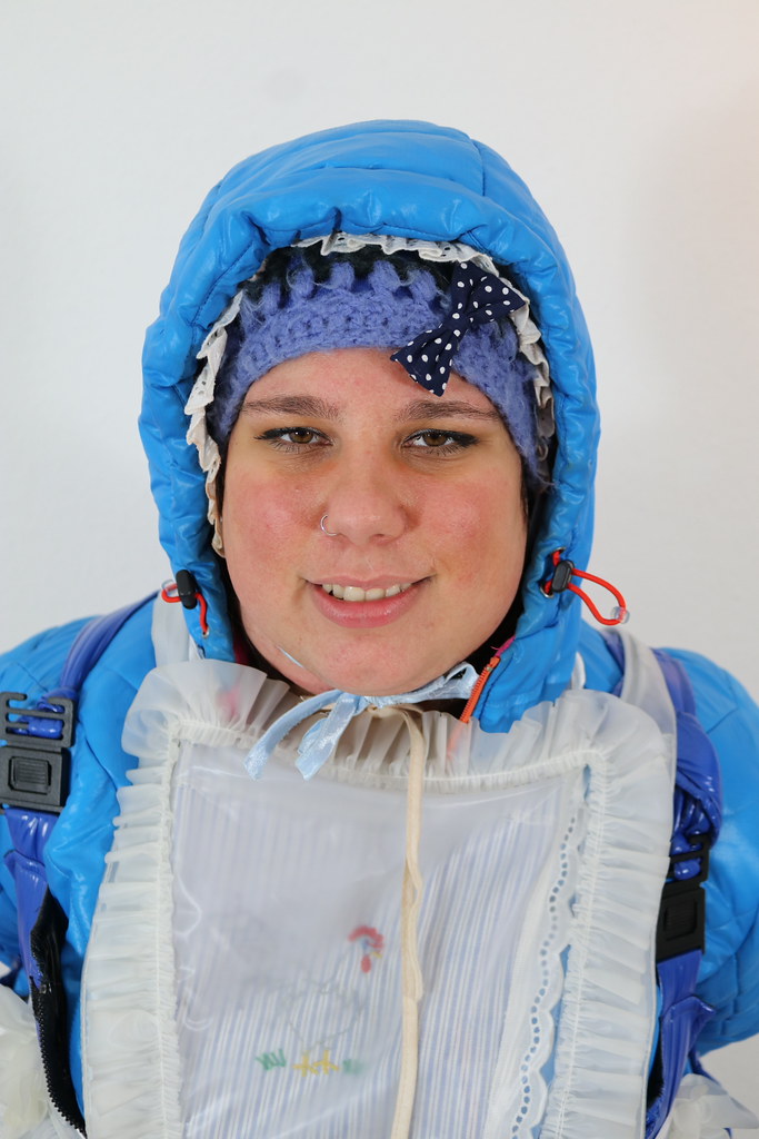 Studio portrait of a person in a bright blue winter parka with hood, lavender knit cap with polka-dot bow, and lace-trim overlay against a white backdrop