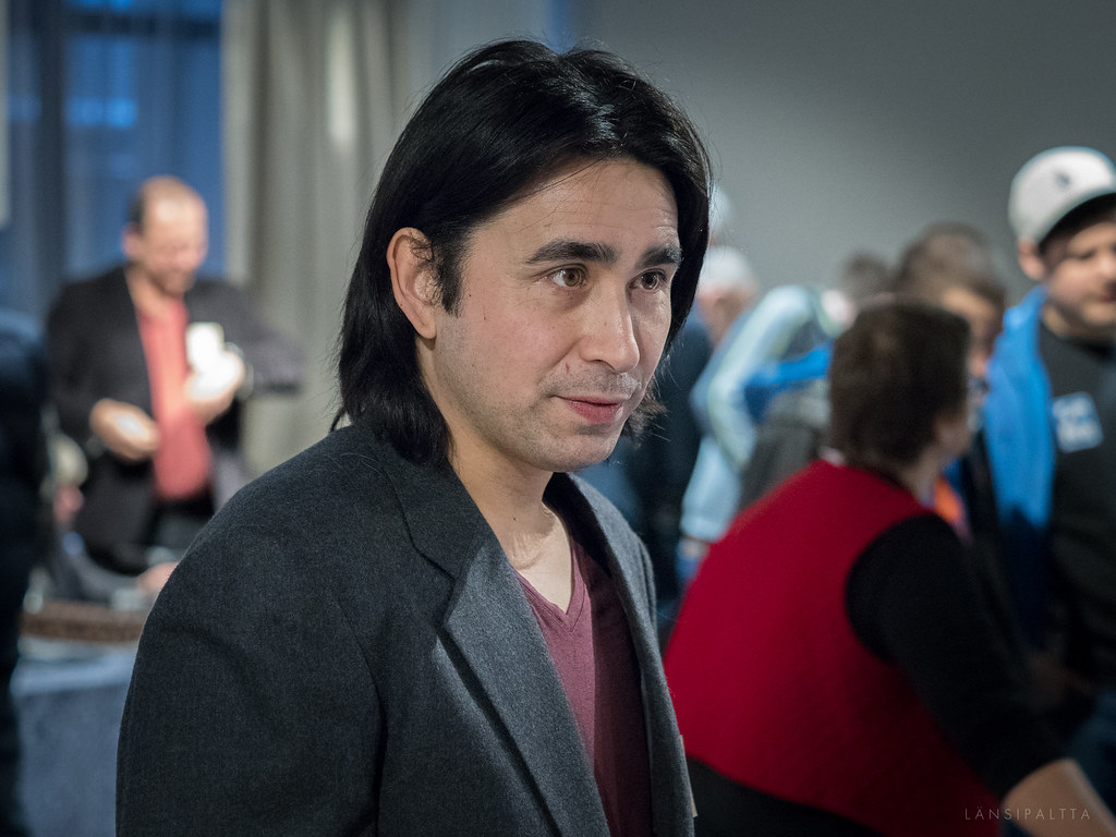 Portrait at a crowded indoor gathering: man with shoulder-length black hair in a charcoal blazer